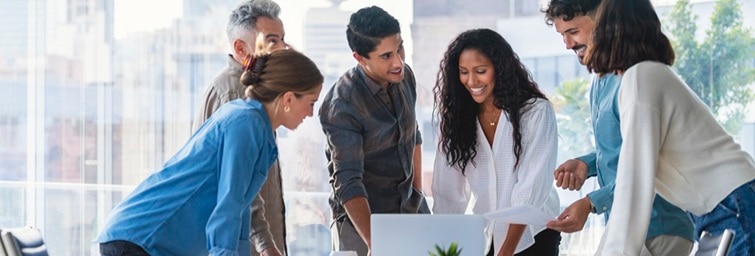 A group of coworkers standing around a table in an office looking down ...